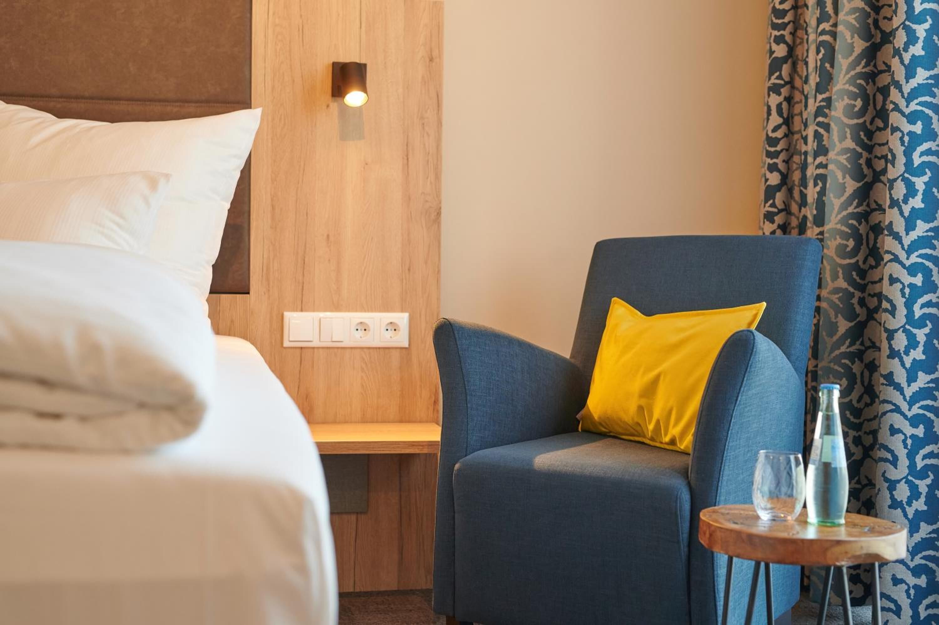Quarto de hotel com uma cama, uma poltrona azul com almofada amarela, uma mesa de madeira pequena com um copo e garrafa, e cortinas estampadas.