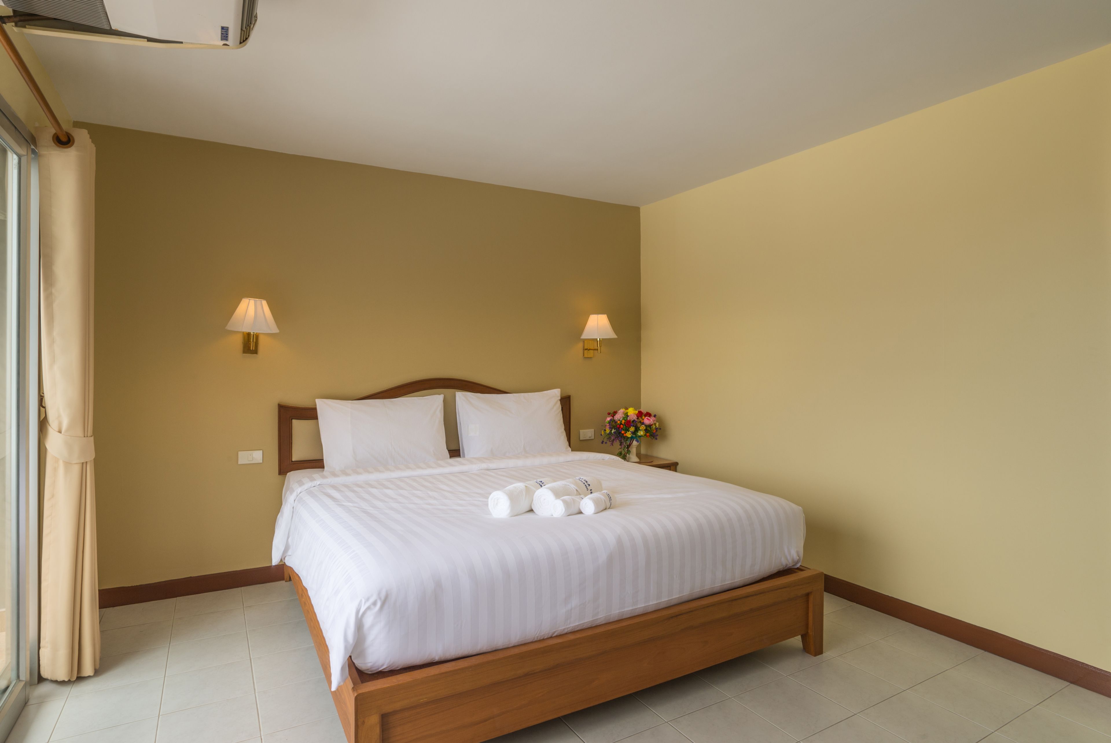 Cama bem arrumada com lençóis brancos e toalhas dobradas, duas luminárias na parede e um vaso com flores em uma mesa de cabeceira.