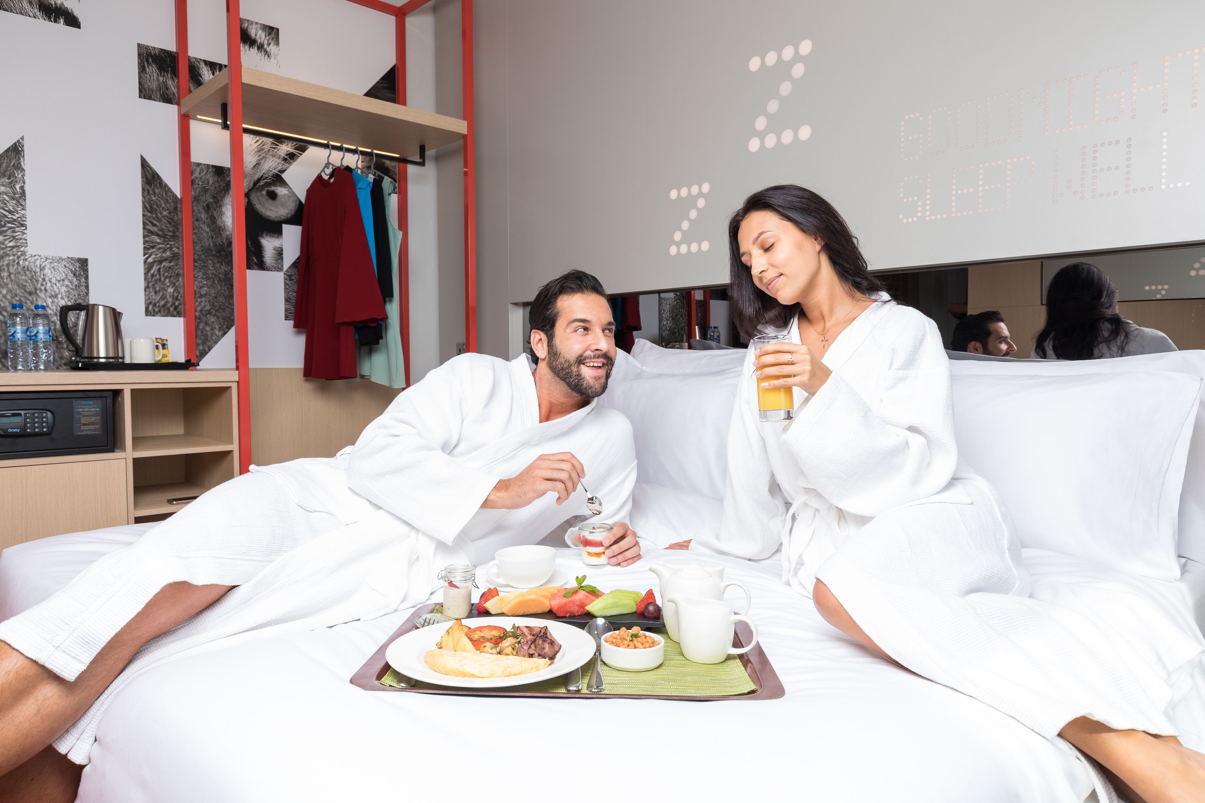Um casal em quarto de hotel moderno, com cama, bandeja de café da manhã e um relógio digital na parede.