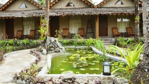 Hotel no estilo de bungalow com telhado de palha, um pequeno lago com nenúfares e um jardim com diversas plantas.