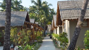 Uma trilha leva a edifícios de estilo tradicional com telhados de palha, cercados por palmeiras e plantas tropicais.