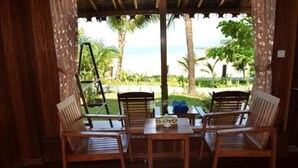 Um deck de madeira com duas cadeiras e uma pequena mesa, com vista para uma praia com palmeiras e o oceano.