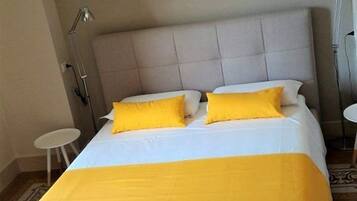 Quarto de hotel com uma cama grande, duas luminárias de cabeceira, uma mesa redonda pequena e uma janela ampla.