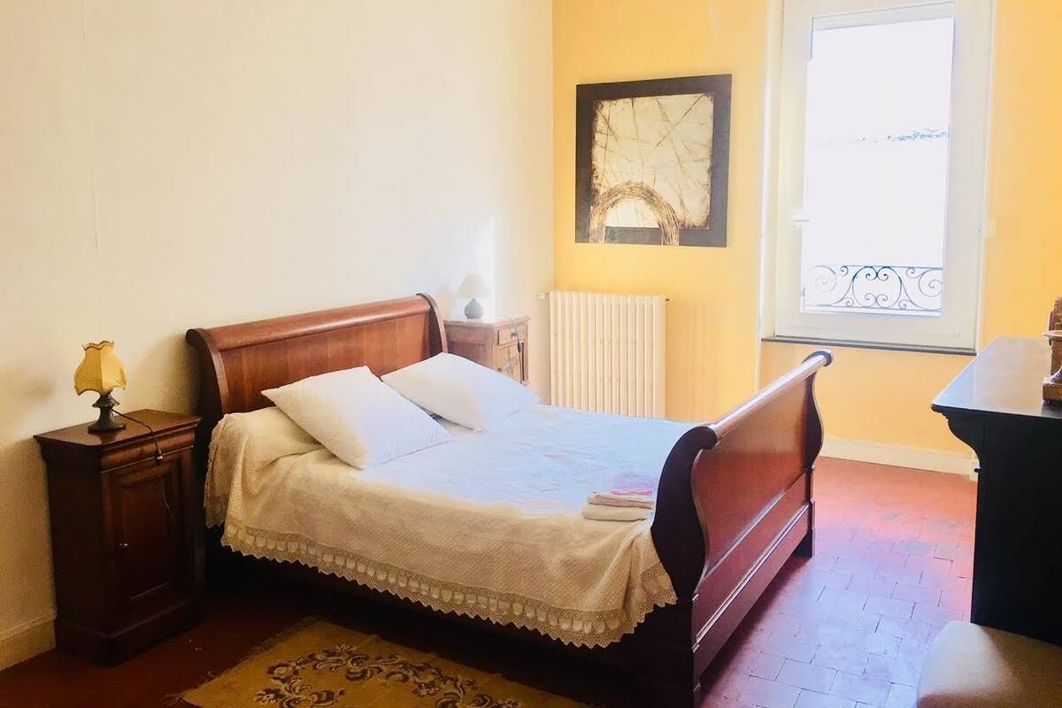 Quarto com cama de madeira, mesa de cabeceira, abajur, radiador e janela com vista.