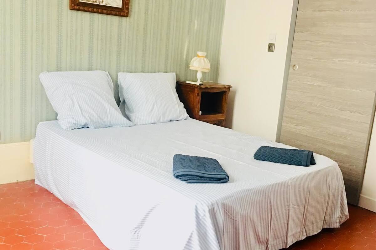 Cama bem arrumada com colcha branca e duas almofadas, uma pequena mesa de cabeceira com abajur, e uma porta de madeira.