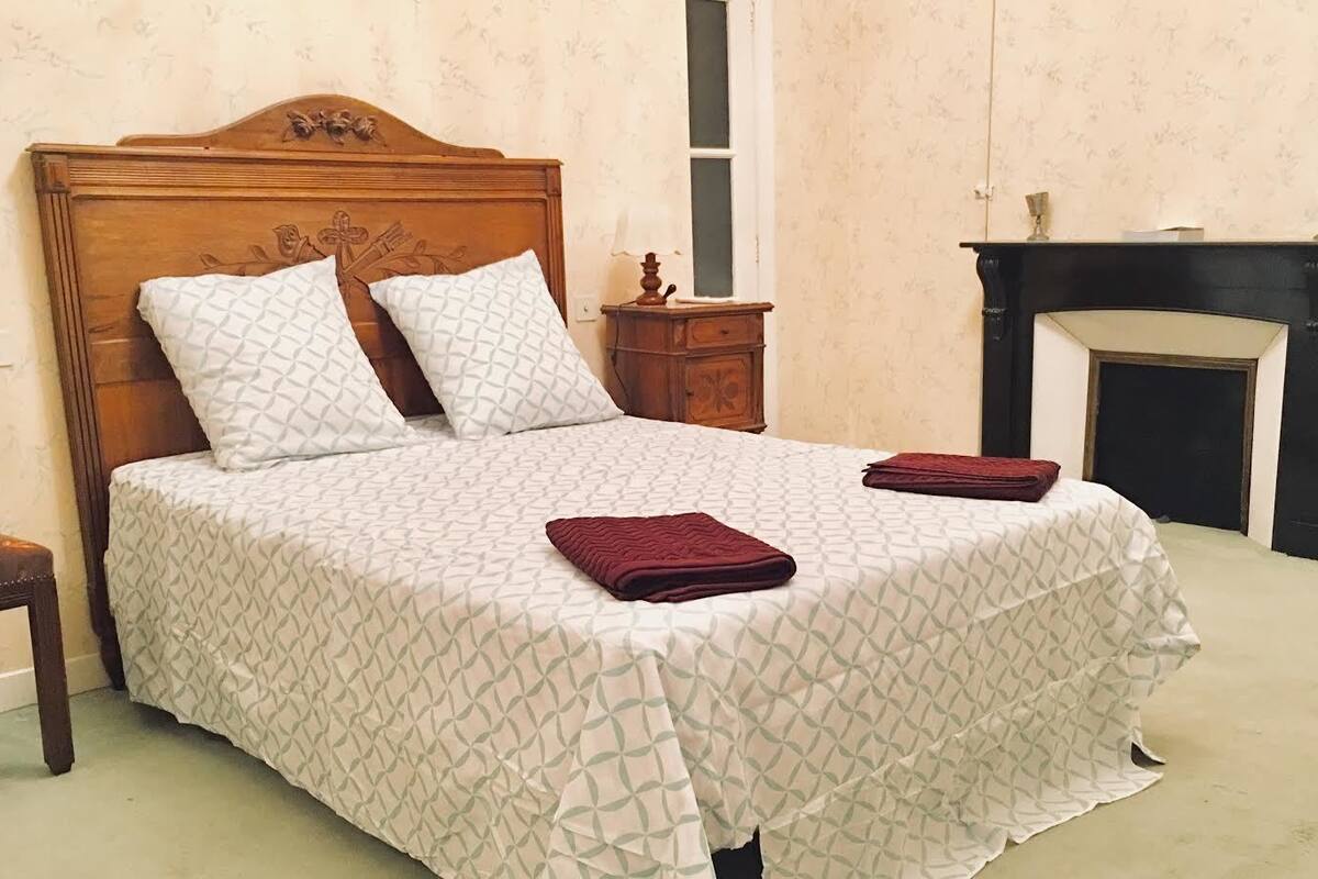 Uma cama bem arrumada com colcha estampada e almofadas, uma pequena mesa de cabeceira de madeira, um cobertor vermelho e uma lareira ao fundo.