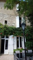 Um edifício de pedra com persianas brancas, uma porta de vidro e uma mesa com cadeiras lá fora.