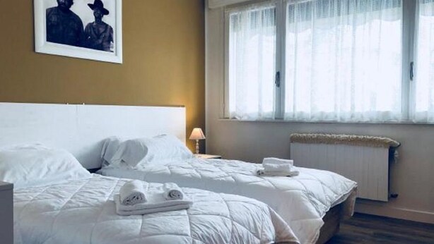 Quarto de hotel com duas camas, uma moldura na parede, uma janela com cortinas translúcidas e um radiador abaixo da janela.