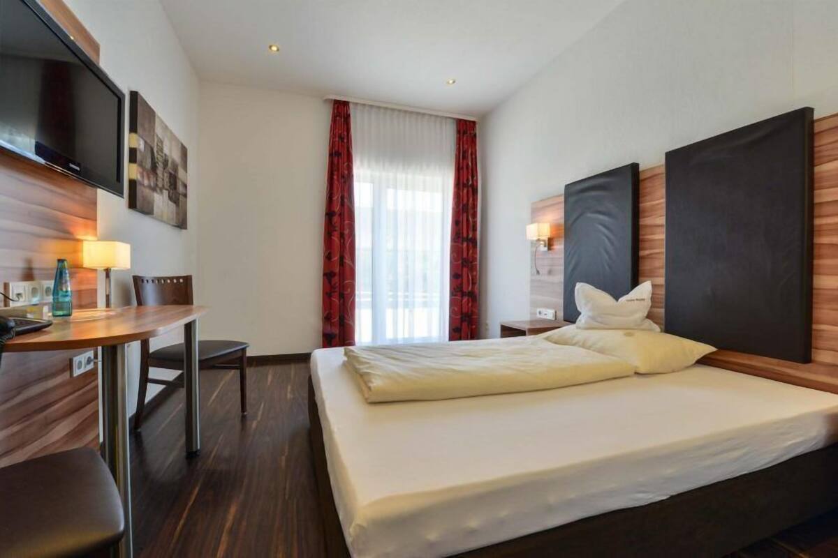Quarto de hotel com cama, escrivaninha com cadeira, televisão fixada na parede e janela com cortinas.