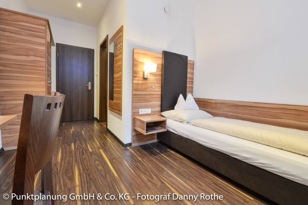Quarto de hotel com chão de madeira, cama com roupa de cama branca, mesa de cabeceira, luminária na parede e um espelho.