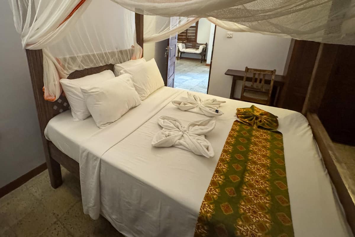 Uma cama bem arrumada com roupa de cama branca e um edredom verde estampado, um dossel branco e uma cadeira ao fundo.