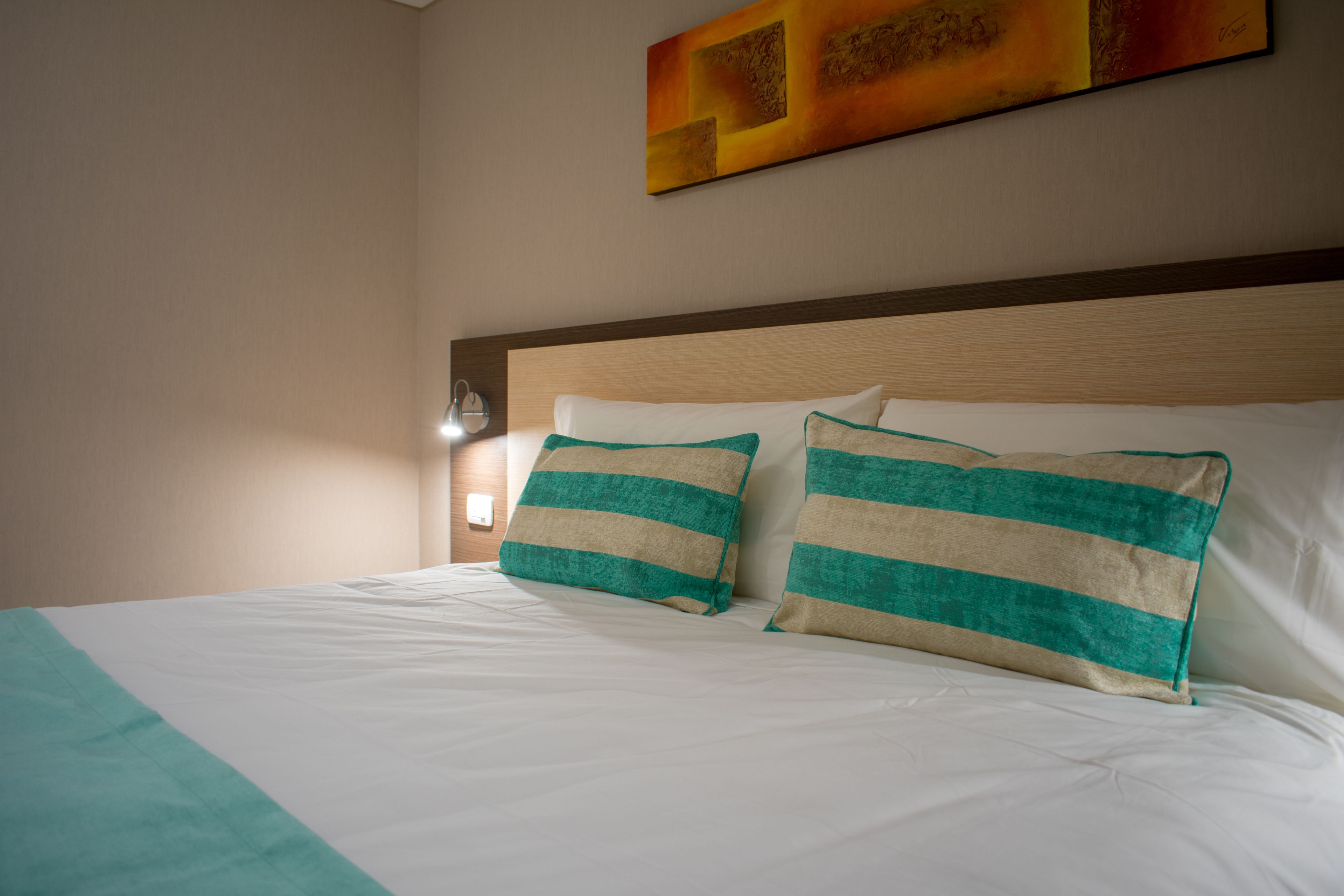 Quarto de hotel com cama bem arrumada, almofadas listradas, um quadro na parede e uma luminária de cabeceira.