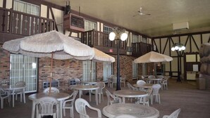 Um espaço de refeições interno com mesas e cadeiras redondas, guarda-sóis de palha e uma parede de tijolos.