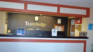Um balcão de recepção com o nome 'Travelodge' em destaque. Há placas informativas e um relógio acima do balcão.