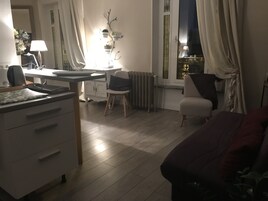 Quarto bem iluminado com escrivaninha, cadeira e cama. Há um radiador e uma janela com cortinas.