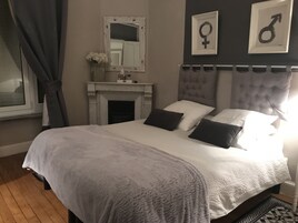 Quarto com uma cama grande, lareira e espelho acima. Há dois símbolos emoldurados na parede.