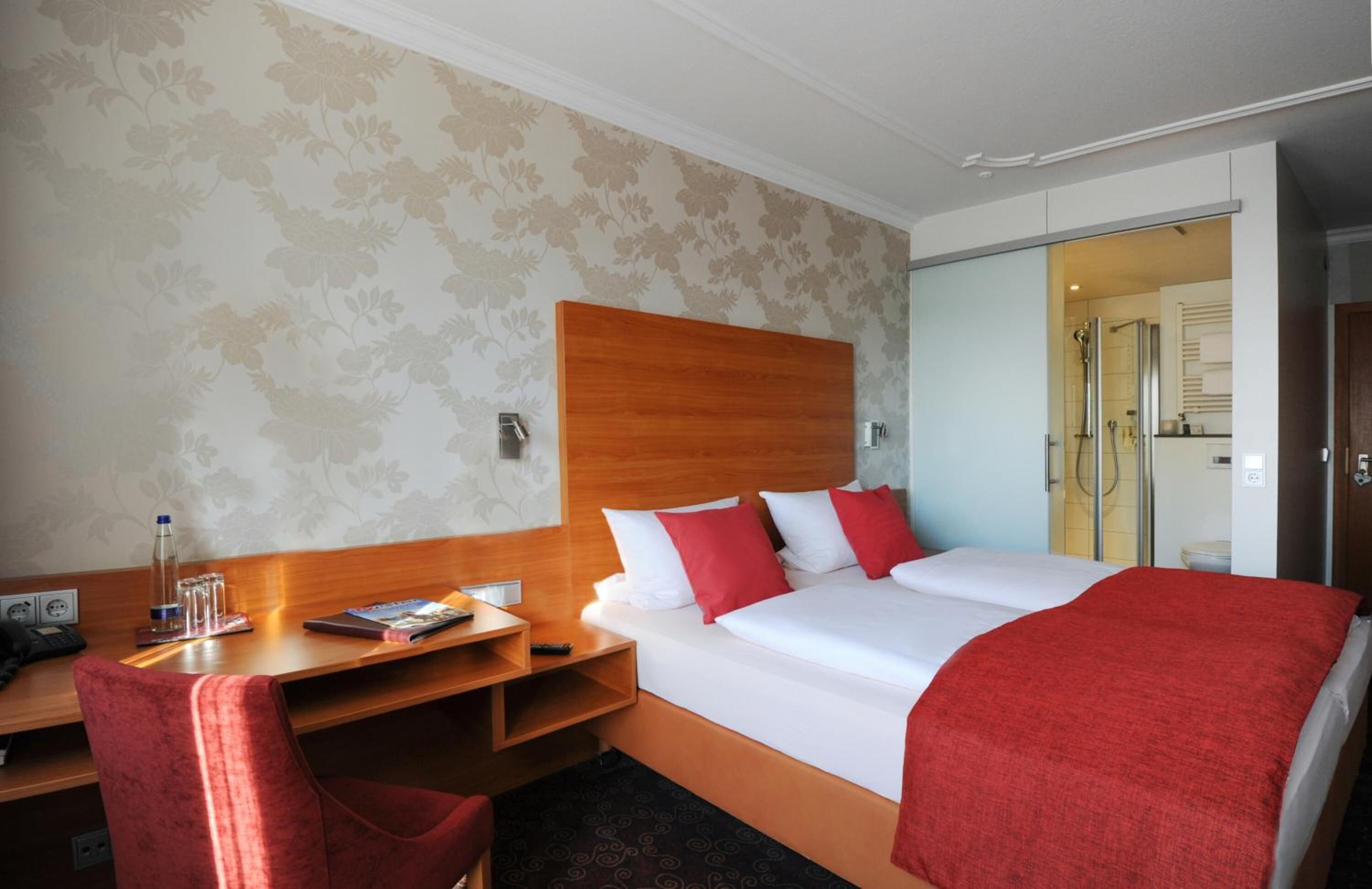 Quarto de hotel com uma cama grande, cabeceira de madeira, escrivaninha com cadeira e área de chuveiro visível através de uma porta de vidro.
