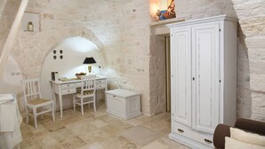 Quarto com parede de pedra, escrivaninha branca com luminária, cadeira, armário e sofá marrom.