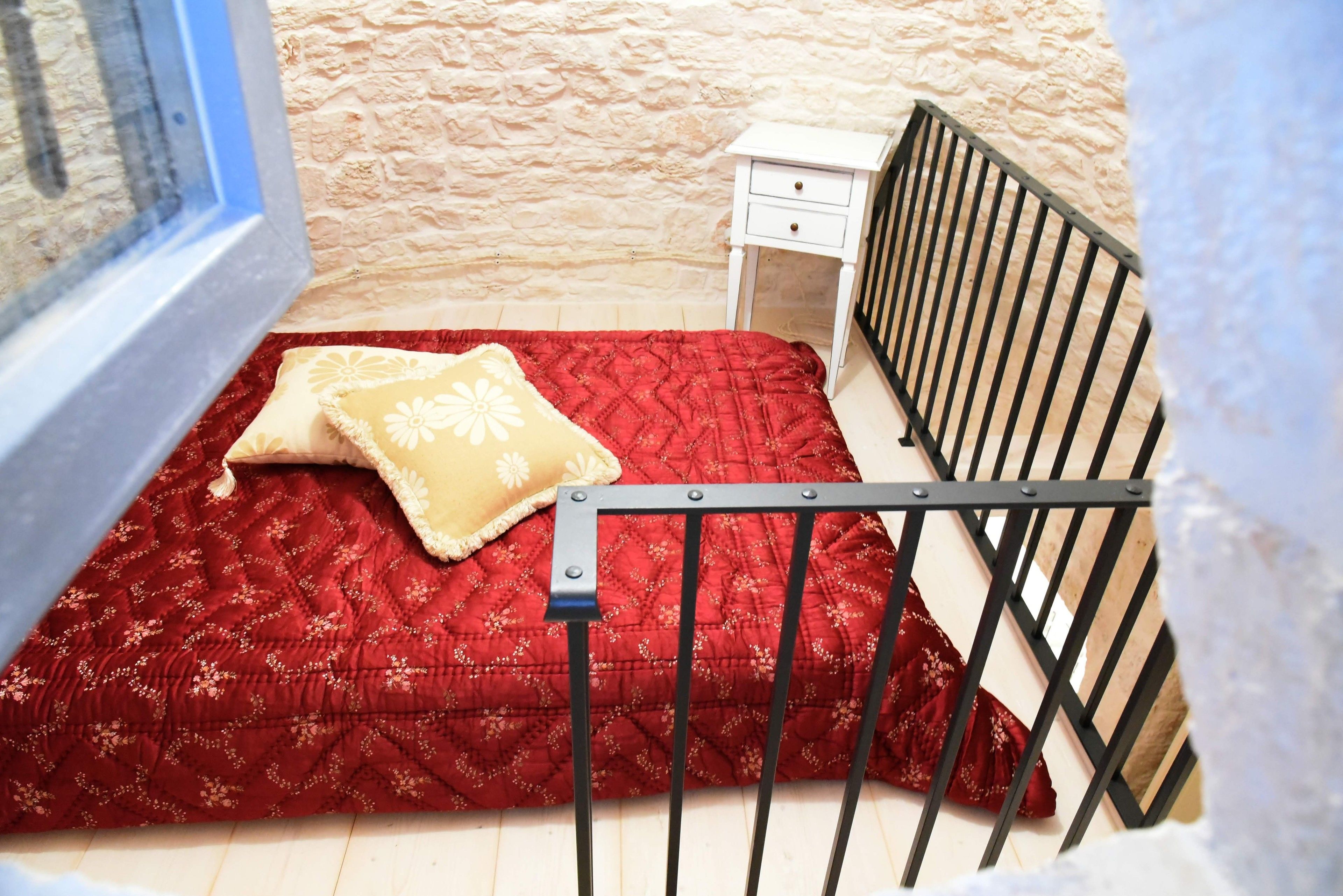Quarto com colcha vermelha, travesseiro bege, mesa de cabeceira branca e grade de cama de metal preto.