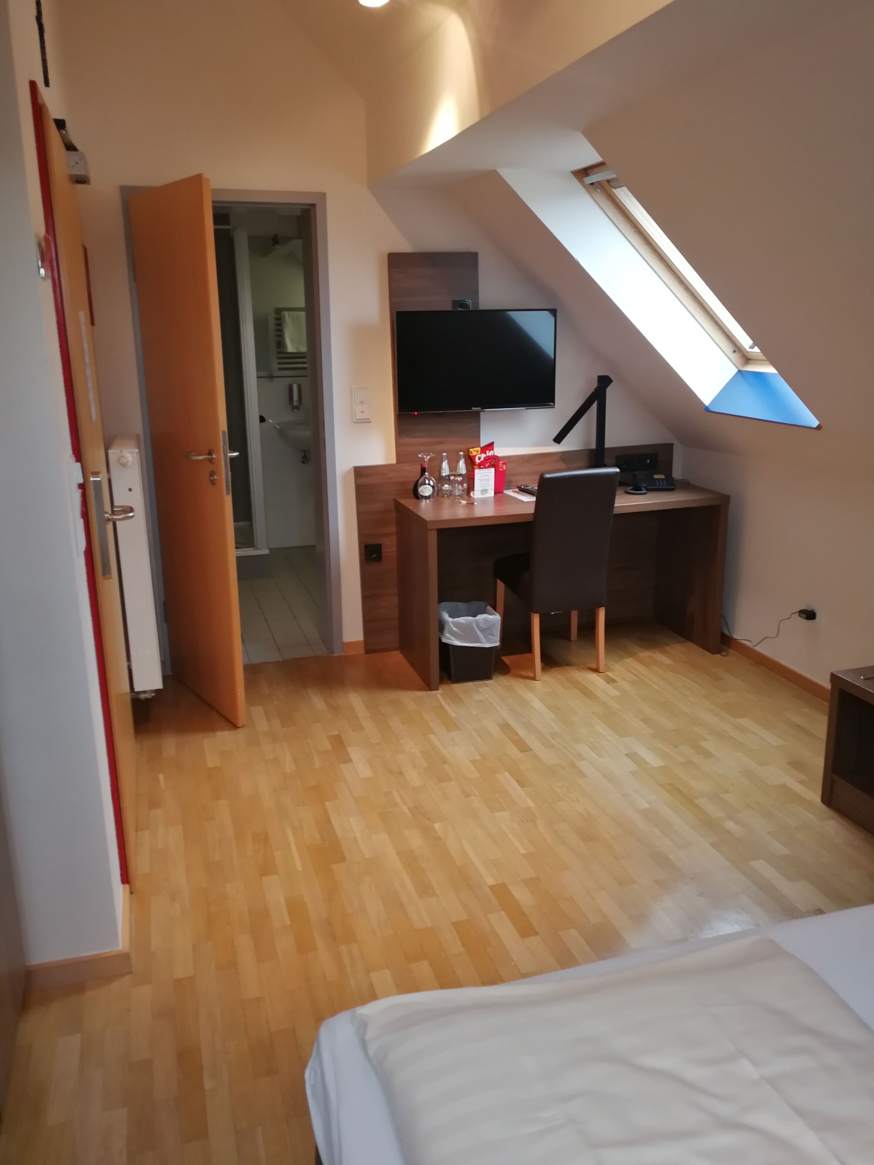 Quarto com piso de madeira, uma cama, uma escrivaninha com cadeira, TV montada na parede e um claraboia.