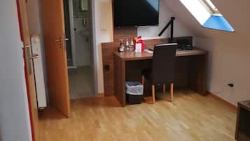 Quarto com piso de madeira, uma cama, uma escrivaninha com cadeira, TV montada na parede e um claraboia.