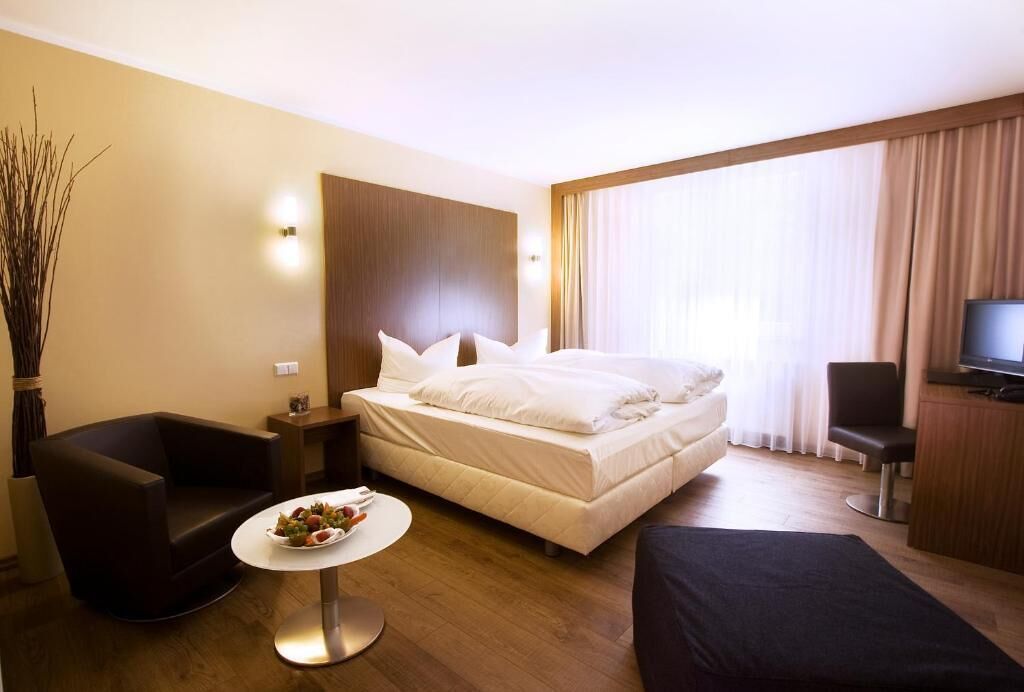 Quarto de hotel com cama, mesa redonda pequena com uma tigela de frutas, sofá, televisão e uma janela grande com cortinas.