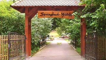 Uma arcada de madeira com uma placa que diz "Springbach-Mühle" dando acesso a um caminho pavimentado.