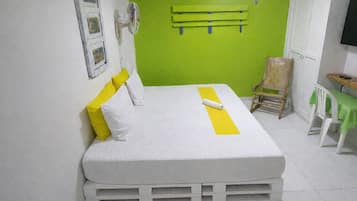 Quarto com uma cama, um ventilador, parede verde e uma pequena mesa com cadeira.