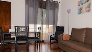 Uma sala de estar com um sofá marrom, duas cadeiras pretas, uma mesa de jantar com cadeiras, um abajur roxo e uma janela com cortinas leves.