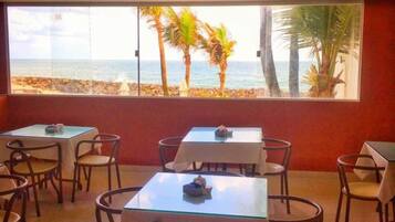 Um restaurante com mesas e cadeiras, vista para a praia e palmeiras.