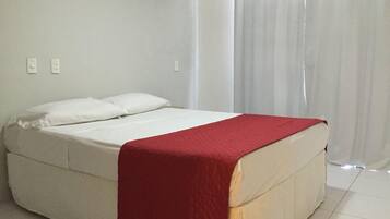 Quarto individual com cama de solteiro, colchão branco, cobertor vermelho e cortinas brancas.