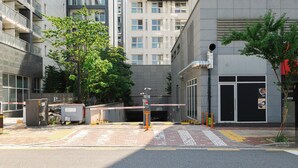 Entrada gradeada para um edifício, com um caminho bem definido, uma árvore e uma lixeira.