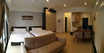 Quarto de hotel com duas camas, um sofá, uma mesa de jantar com cadeiras, uma cozinha e uma janela grande com cortinas.