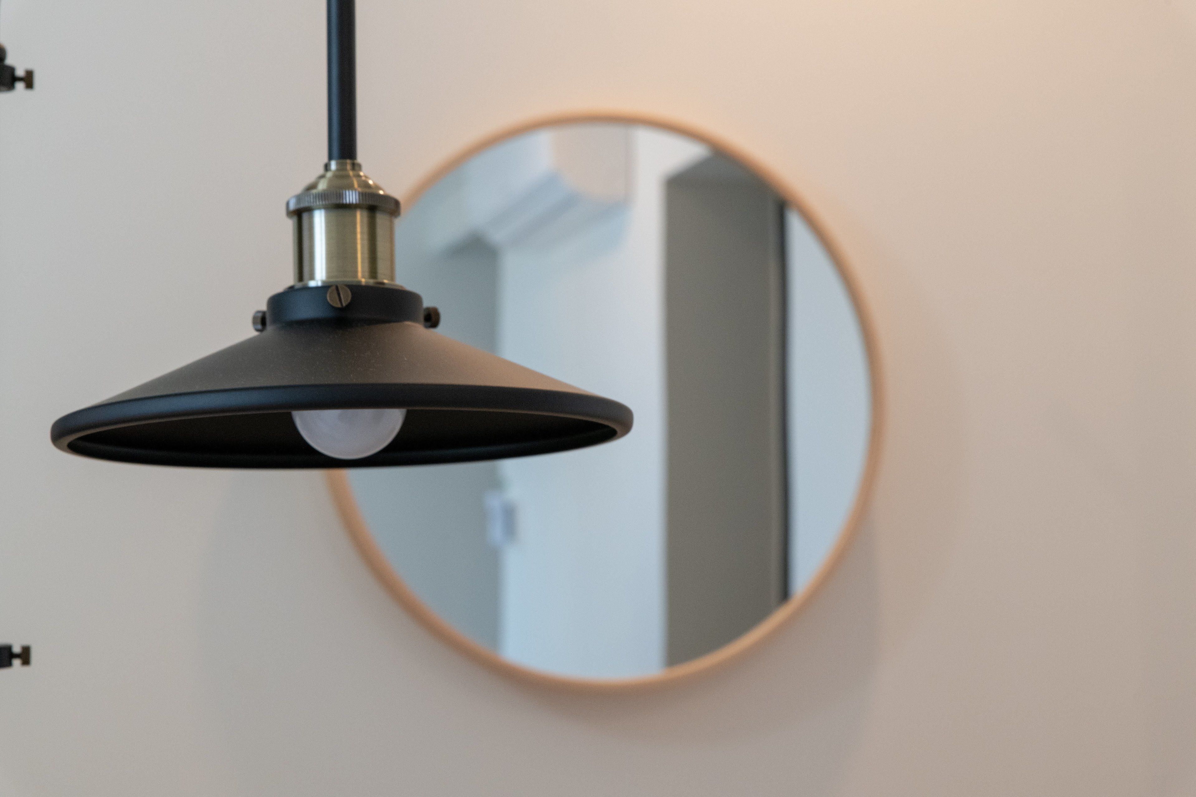Luminária pendente com cúpula preta e base metálica, fixada em parede com espelho redondo refletindo uma porta.