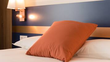 Cama bem arrumada com almofadas brancas e laranja, luminária fixada na parede e esquema de cores azul e dourado.