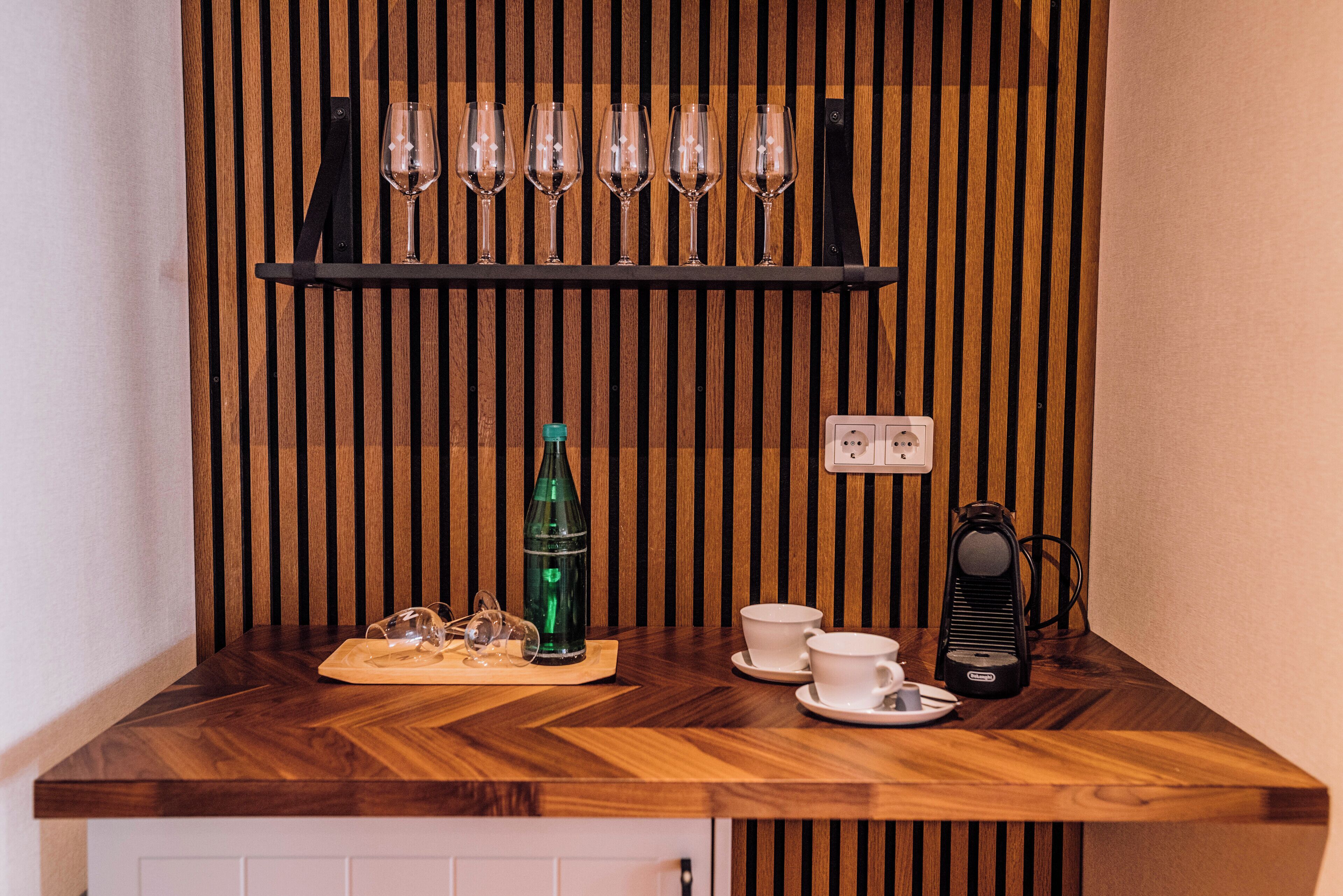 Uma área de bar com balcão de madeira, prateleira com copos de vinho, uma garrafa verde e uma máquina de café.