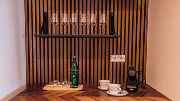 Uma área de bar com balcão de madeira, prateleira com copos de vinho, uma garrafa verde e uma máquina de café.