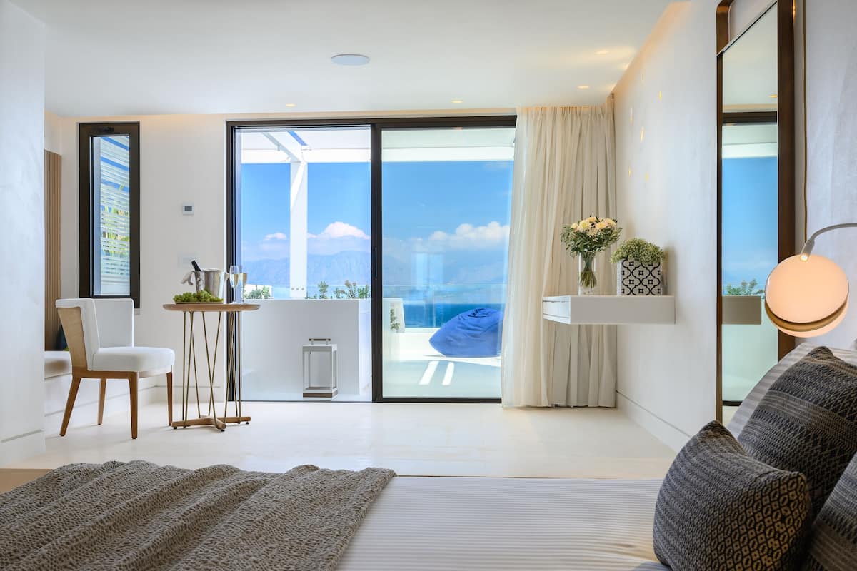 Quarto de hotel moderno com uma grande porta de vidro deslizante que dá para uma varanda com vista para o mar e montanhas.
