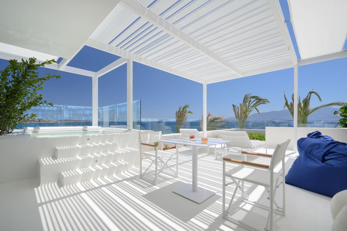 Uma área de estar moderna ao ar livre, com móveis brancos, uma mesa com bebidas e vista para o oceano e a paisagem urbana.