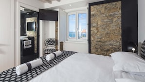 Quarto com cama, parede de pedra decorativa, vista para o mar, banheiro com pia e uma cadeira.