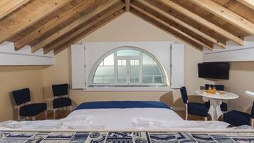 Quarto com teto de madeira, uma cama grande, uma janela redonda, uma TV e uma pequena mesa com cadeiras.