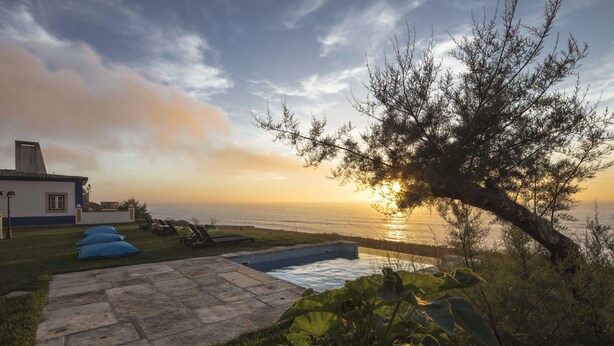 Área de piscina com vista para o pôr do sol sobre o oceano, uma casa e uma árvore.
