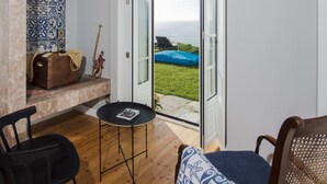 Um quarto com piso de madeira, lareira, uma mesa, uma cadeira e vista para o mar por uma porta aberta.