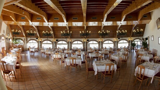 Ampla sala de jantar com alto teto de madeira e várias mesas postas.