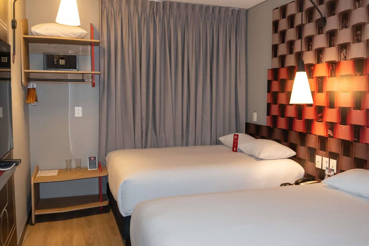 Quarto de hotel com duas camas, um micro-ondas, uma mesa pequena e uma luminária fixada na parede.