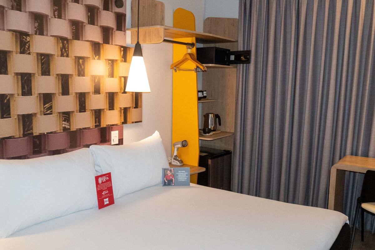 Quarto de hotel com uma cama, candeeiro suspenso, mesa pequena, cadeira, cabideiro e prateleira na parede com uma chaleira.