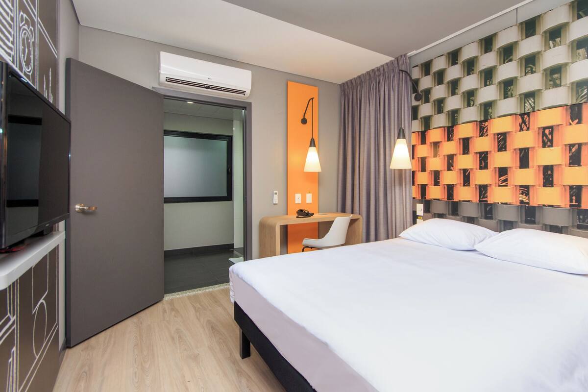 Quarto de hotel com cama, escrivaninha, cadeira, televis ão e ar condicionado.