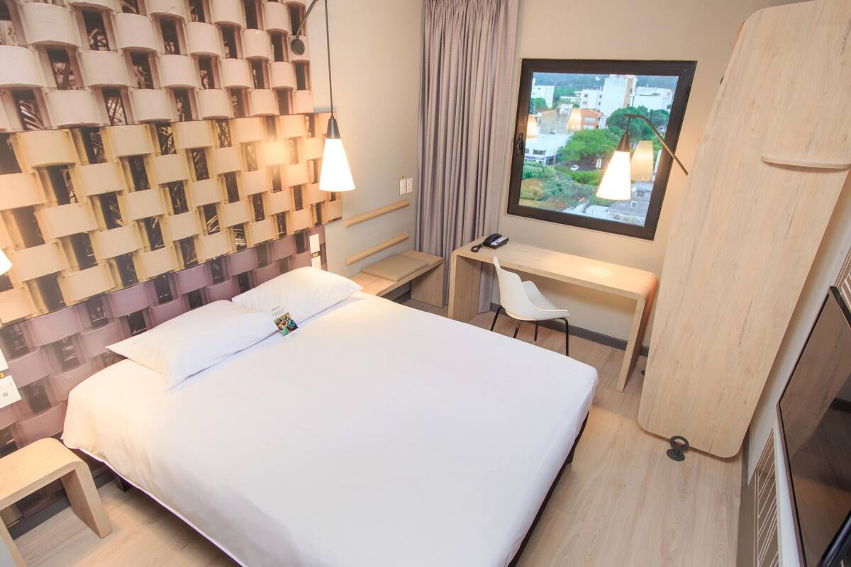Quarto de hotel com cama, escrivaninha, cadeira e vista para a cidade através de uma janela.