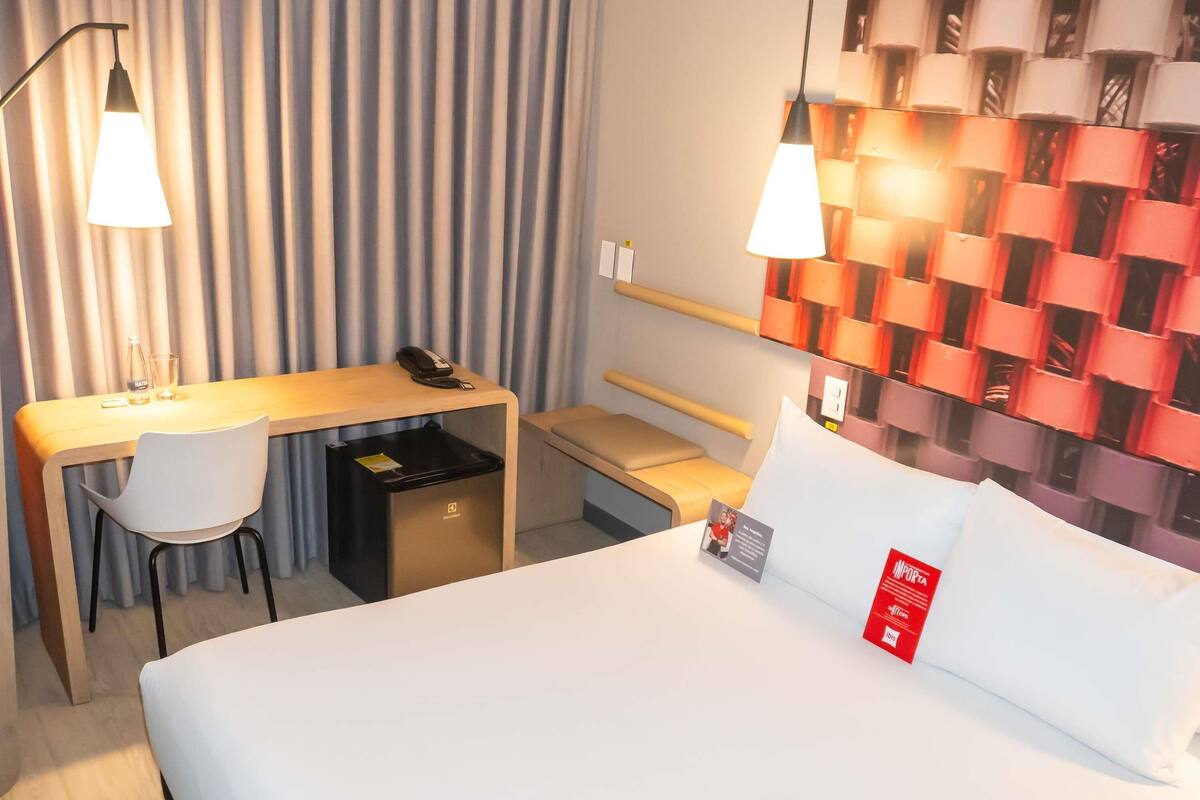 Quarto de hotel com cama, escrivaninha, cadeira, lixeira e luminária de parede.