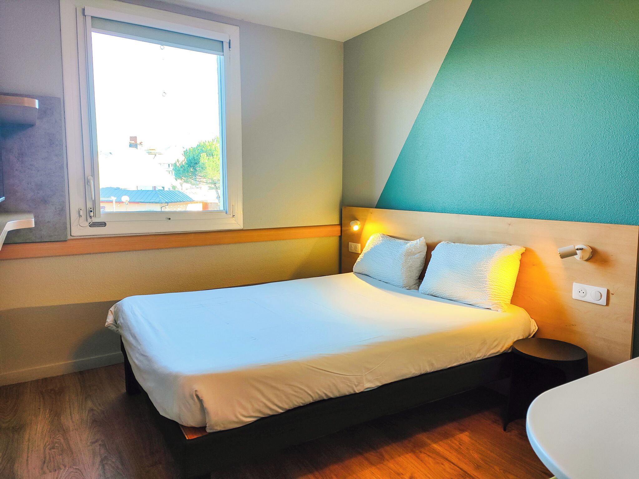 Uma cama bem arrumada com lençóis brancos, uma mesa de cabeceira, um pequeno banquinho e uma janela com vista para prédios e árvores.